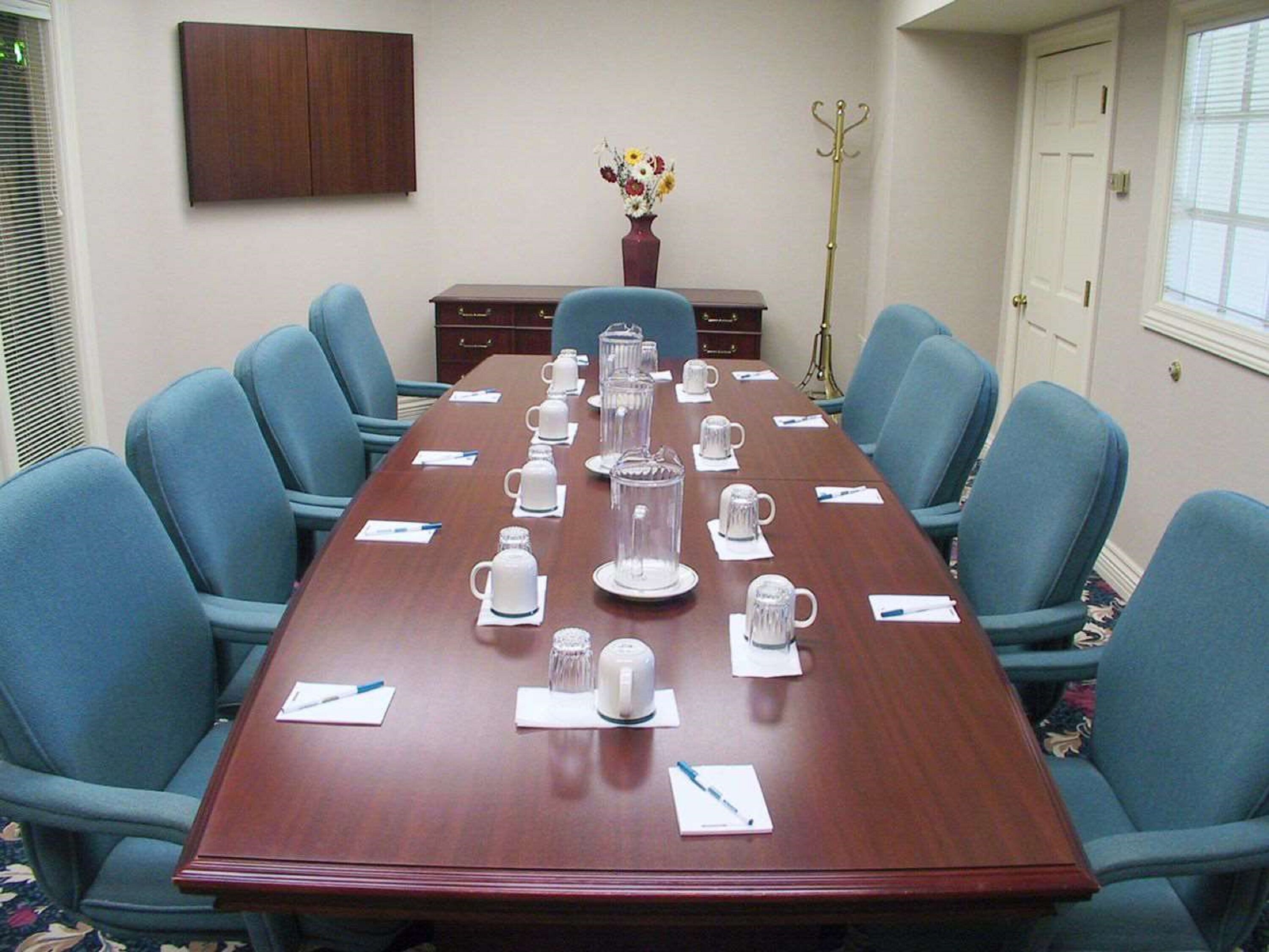 Uma sala de conferências com uma mesa comprida preparada para uma reunião, rodeada por cadeiras azuis, um armário de madeira e um vaso com flores.