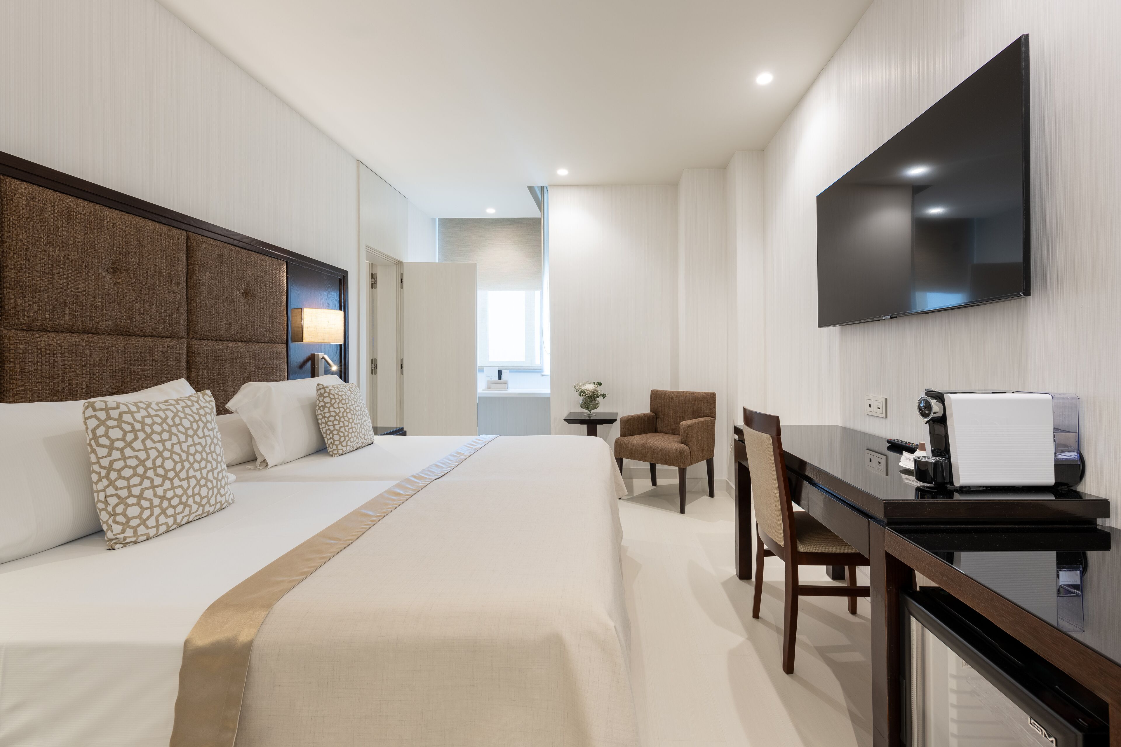 Quarto de hotel com uma cama grande, uma escrivaninha com computador, uma cadeira e uma televisão fixada na parede.