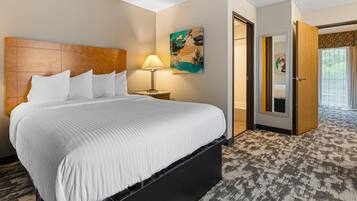 Quarto de hotel com uma cama grande, uma mesa de cabeceira com abajur, um quadro na parede e uma porta que dá para outro cômodo.