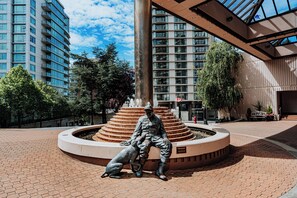 Uma estátua de um homem e um cão numa fonte circular, rodeada por um calçamento e edifícios ao fundo.