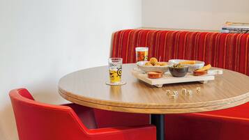 Uma área de refeições com uma mesa redonda, cadeiras vermelhas e um banco. Há um quadro na parede, um tabuleiro com comida e bebidas, e dados na mesa.