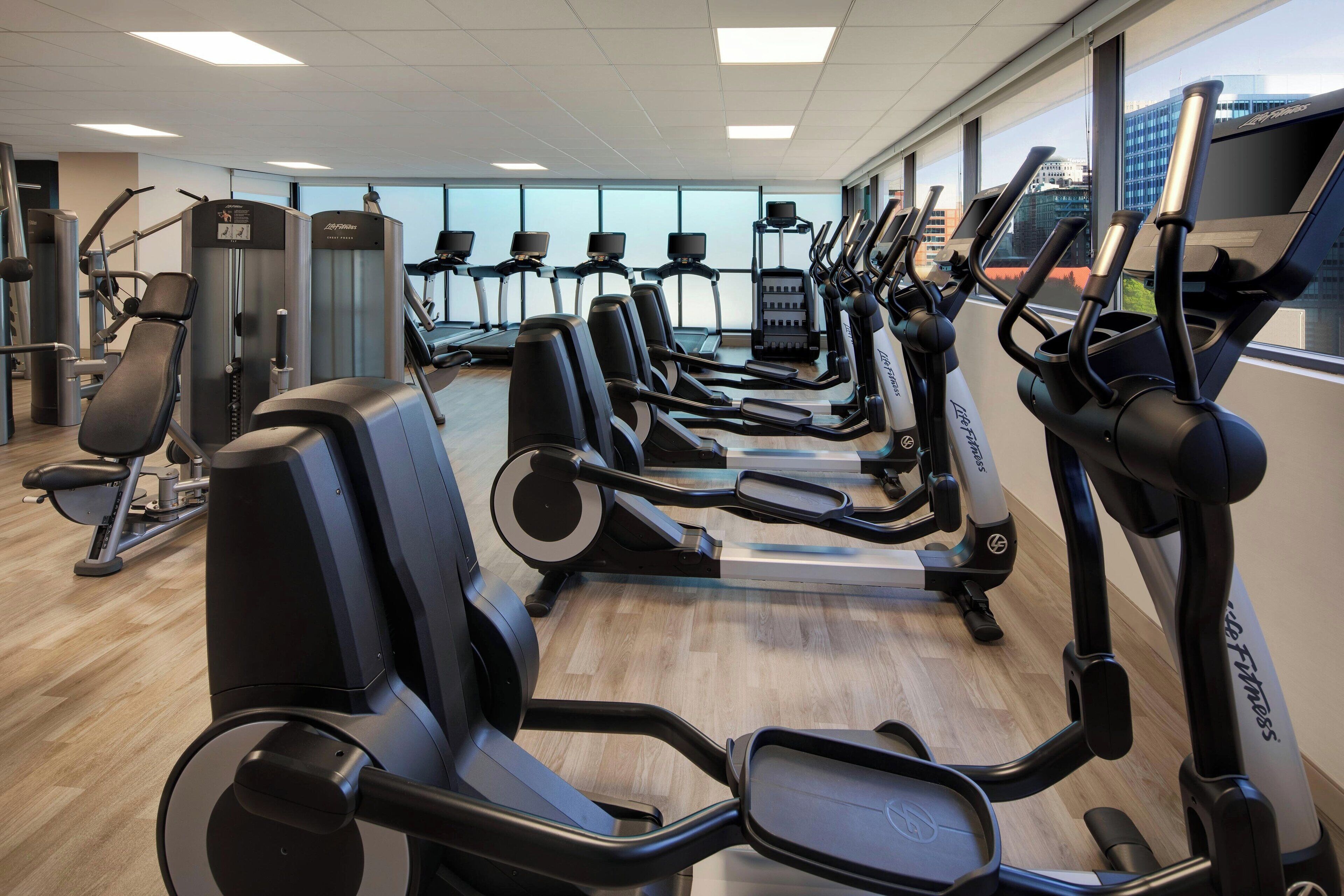 Uma academia com várias esteiras e aparelhos de exercícios, amplas janelas com vista para a cidade e piso de madeira.