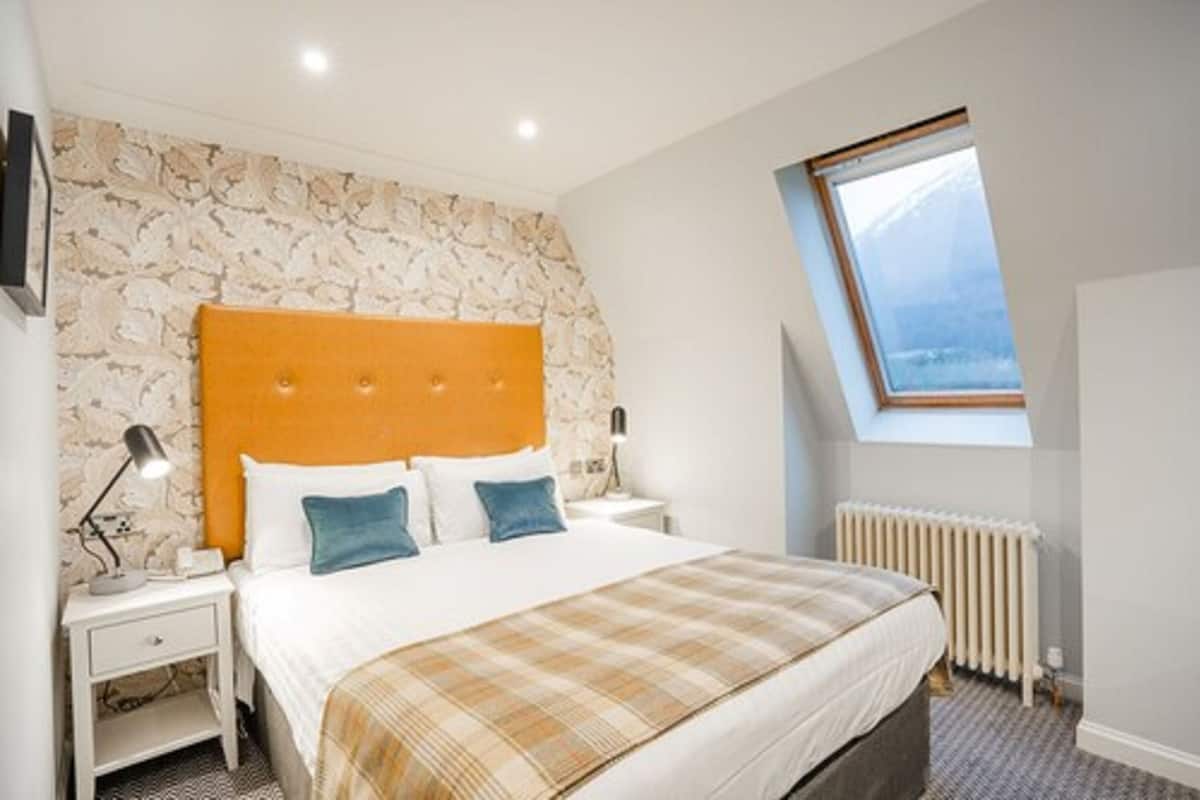 Um quarto com parede de padrão de pedra, uma cama com colcha xadrez, uma mesa de cabeceira com abajur e uma janela com vista para o céu.