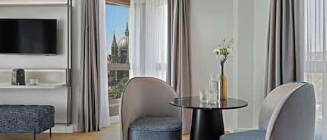 Quarto de hotel moderno com uma janela grande, televisão de tela plana, mesa redonda com cadeiras e um pequeno sofá.