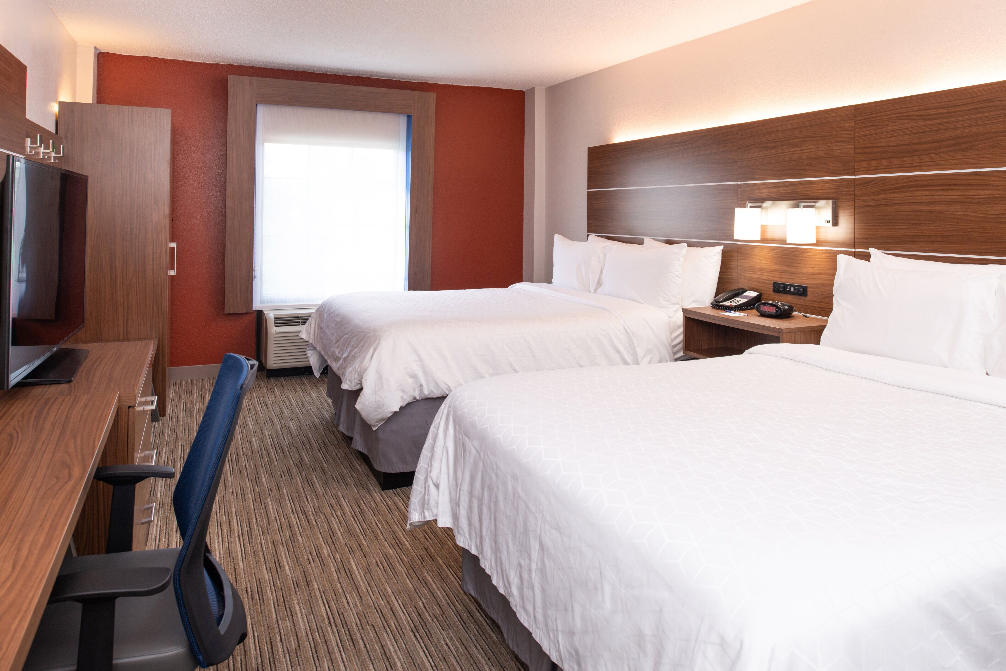 Quarto de hotel com duas camas, uma escrivaninha, uma cadeira, uma televisão e uma janela com persianas.