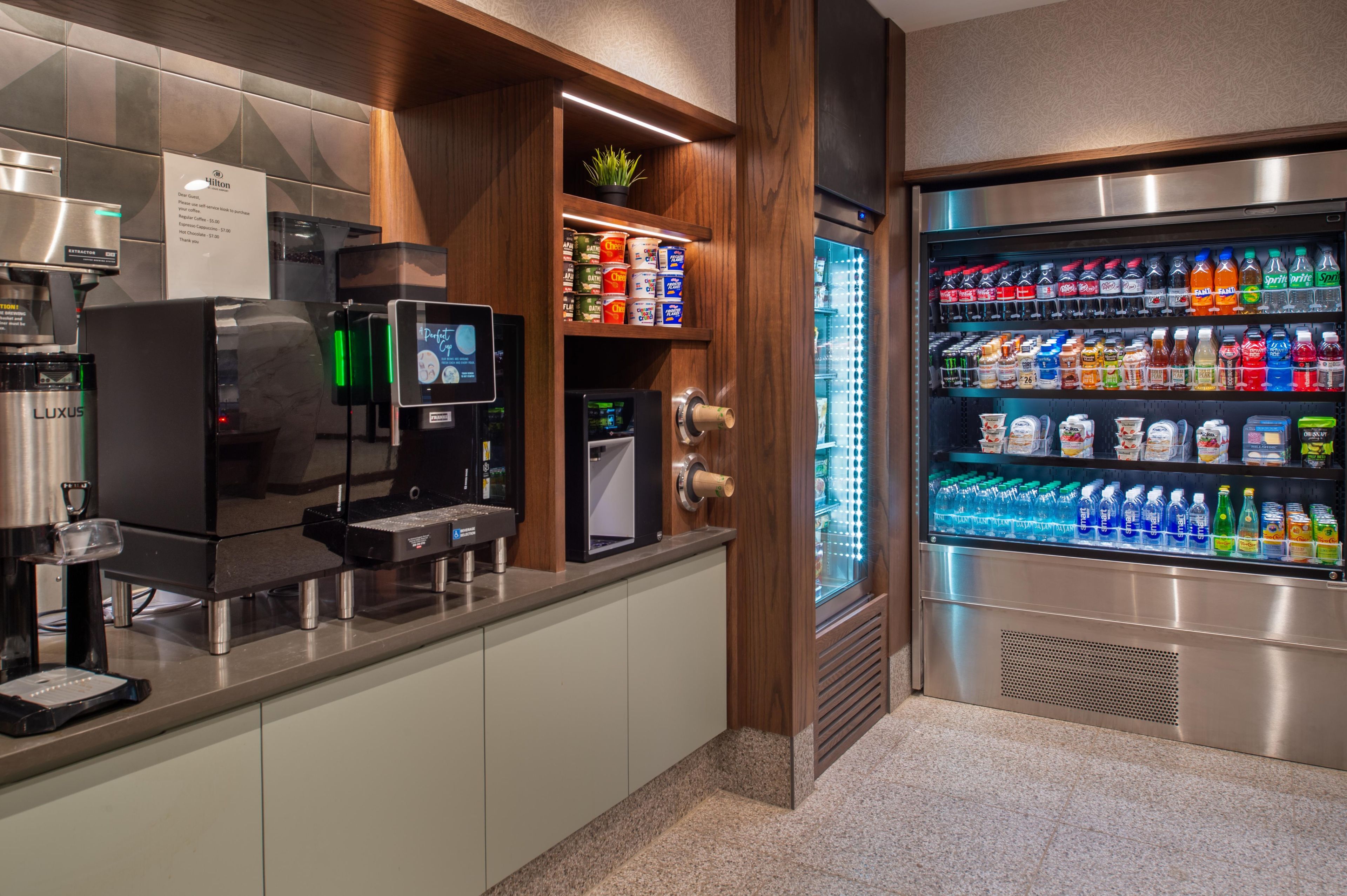Uma estação de café para autosserviço, com máquina de café, terminal de pagamento e refrigerador de bebidas.