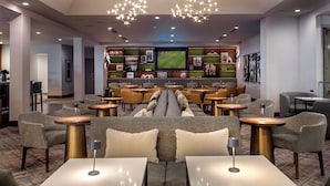Um saguão de hotel com área de estar central, incluindo sofás e mesas, uma grande tela de TV exibindo um jogo esportivo e um bar com cadeiras e balcão.