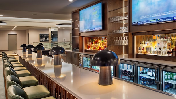 Uma área de bar com um balcão comprido, diversas opções de assento e uma televisão exibindo um programa.
