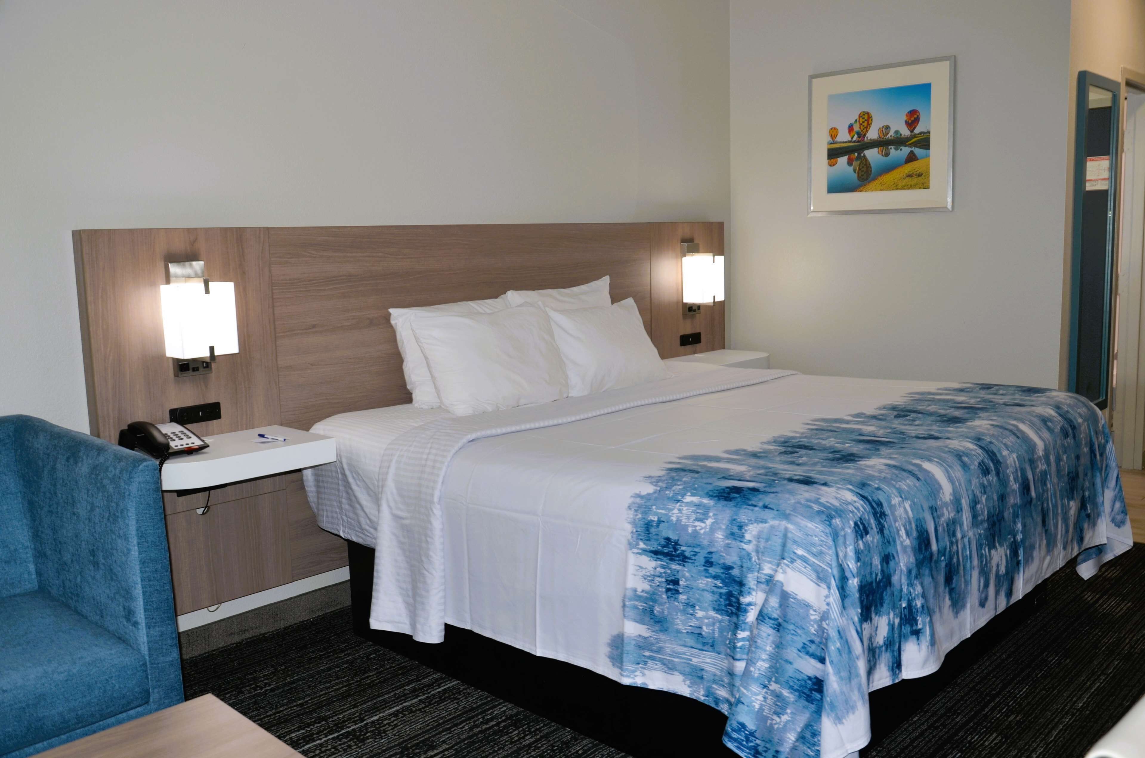 Quarto de hotel com uma cama, mesas de cabeceira, escrivaninha, uma cadeira e um quadro na parede.