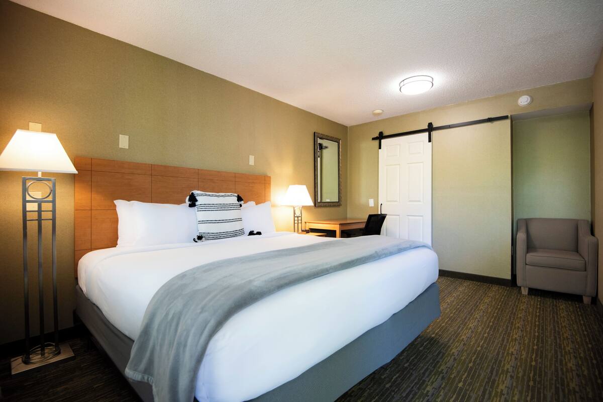 Quarto de hotel com uma cama grande, duas luminárias de cabeceira, uma escrivaninha, uma cadeira e uma porta de correr.