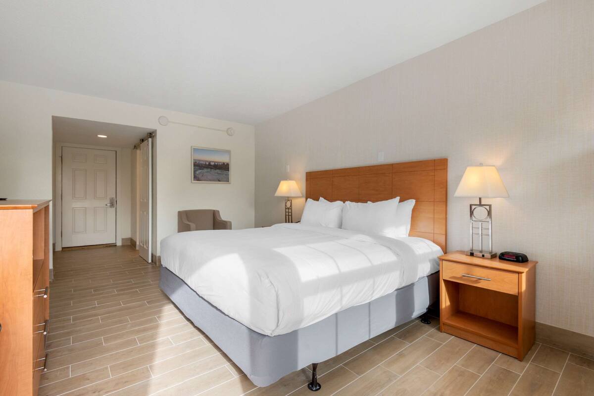 Quarto de hotel com uma cama grande, abajures, uma mesinha de cabeceira de madeira, uma cadeira e uma porta que dá para outro quarto.
