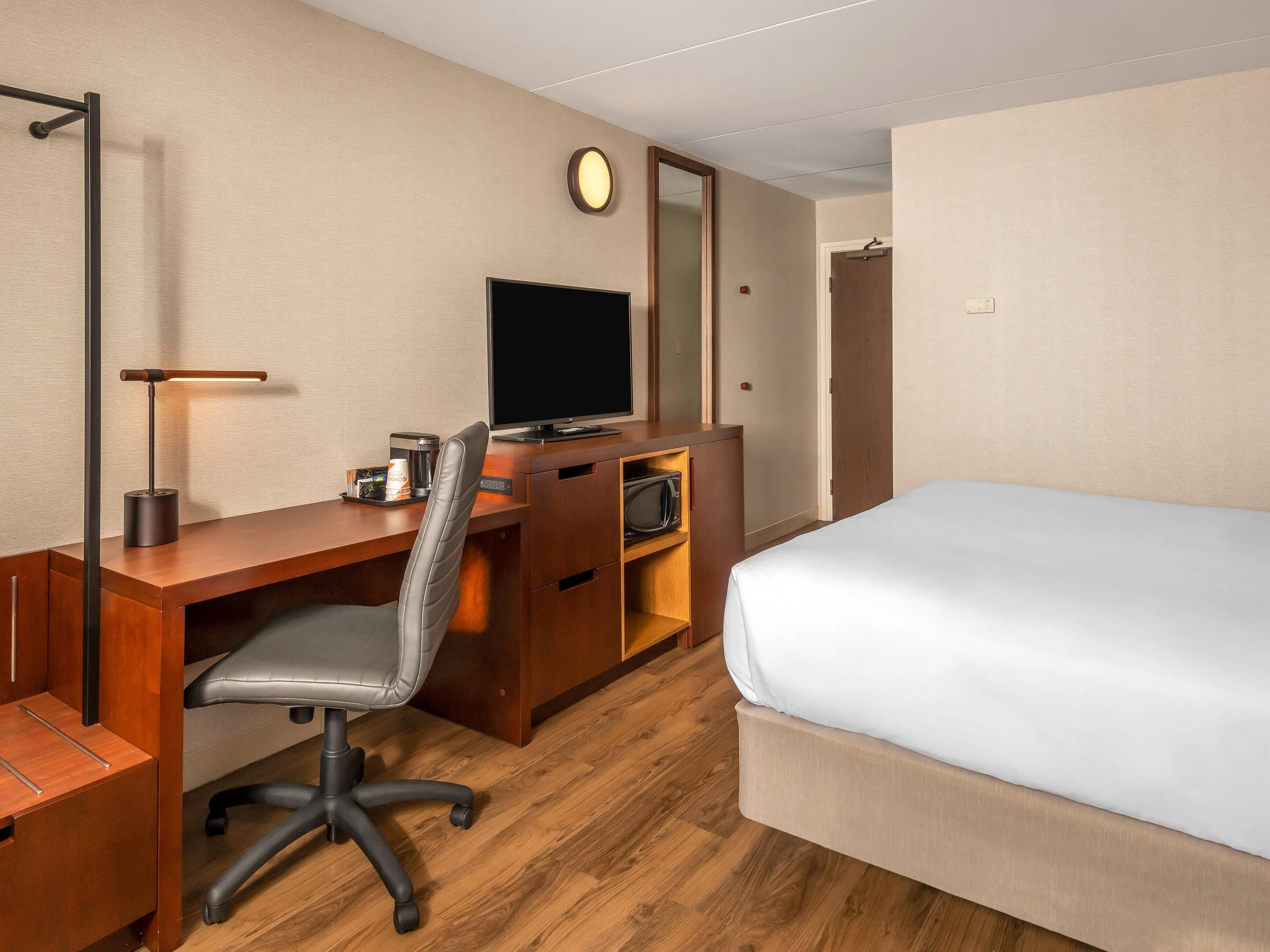 Quarto de hotel com secretária de madeira, televisão, cadeira, cama e micro-ondas.