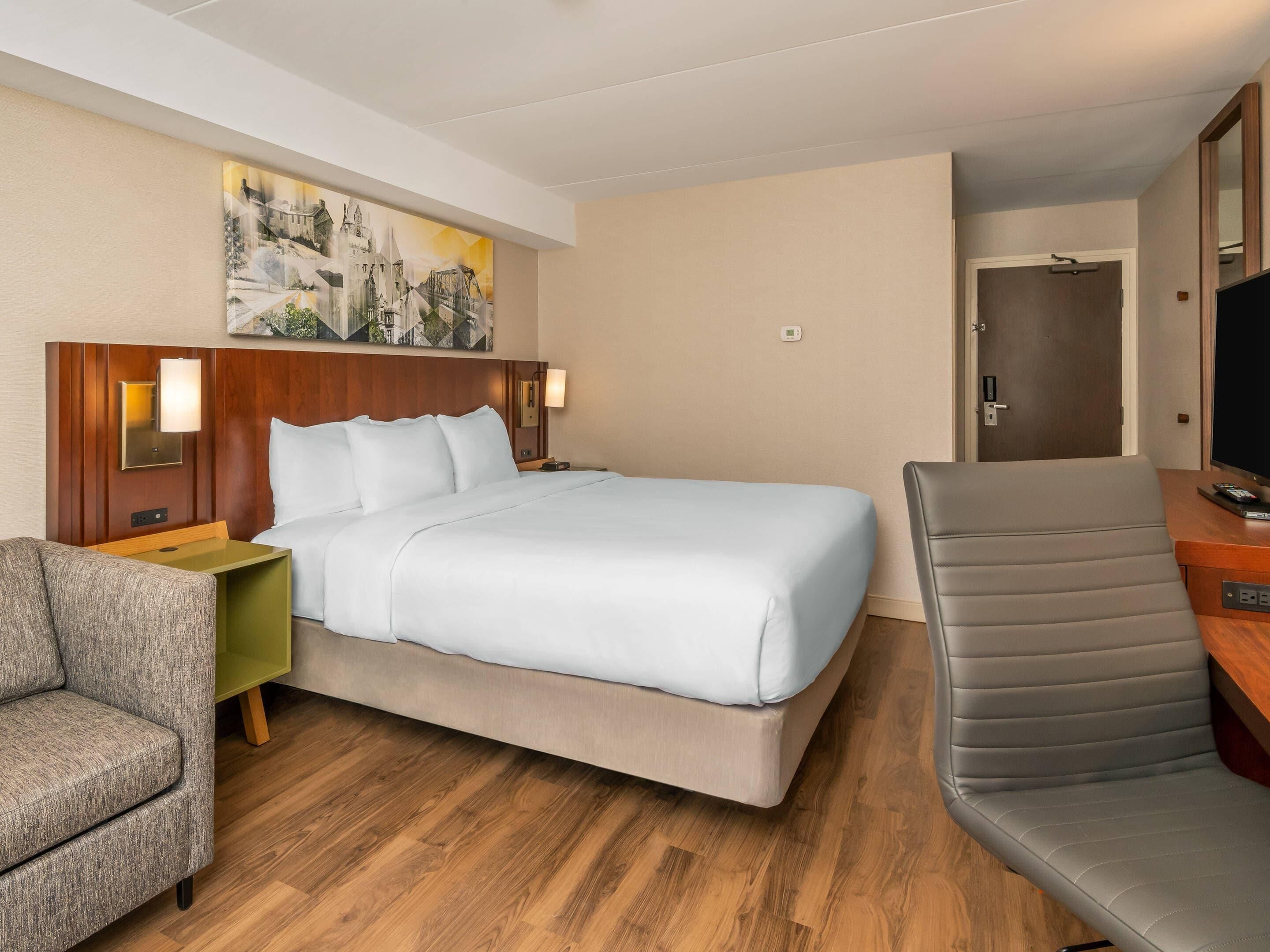 Quarto de hotel com cama, cadeira, escrivaninha, um quadro na parede e uma porta.