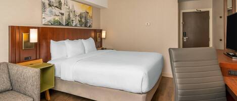 Quarto de hotel com cama, cadeira, escrivaninha, um quadro na parede e uma porta.