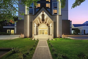 Entrada moderna de hotel com arco bem iluminado, caminho pavimentado e gramado.