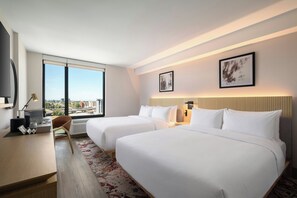 Quarto de hotel com duas camas, uma escrivaninha, uma cadeira, uma televisão e uma janela grande com vista para a cidade.