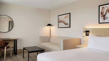 Quarto de hotel com cama, sofá, escrivaninha, cadeira, mesa pequena, abajur e duas quadros na parede.