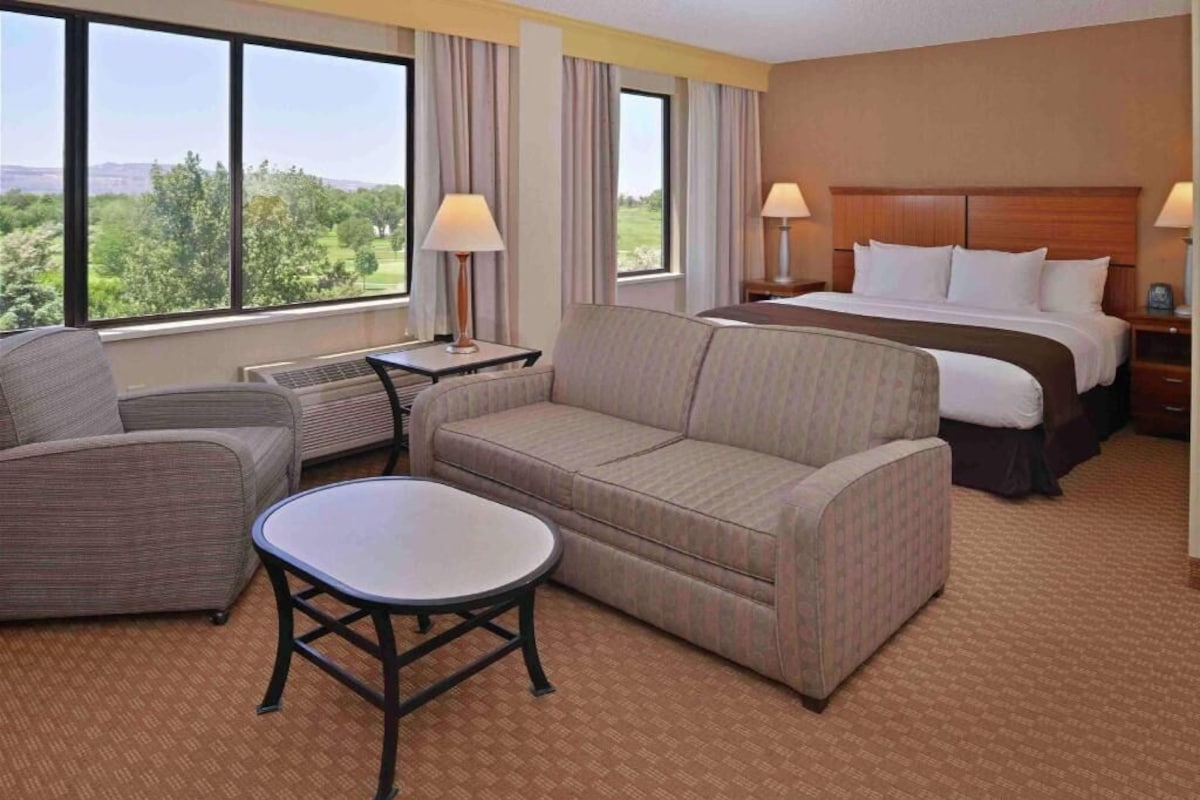 Quarto de hotel com cama, dois sofás, uma mesinha e vista para o verde.