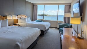 Quarto de hotel com duas camas, uma escrivaninha, uma cadeira e uma janela grande com vista para a água.
