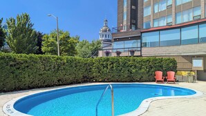 Piscina com escada de metal, cercada por um hedge, e adjacente a um edifício com varanda.