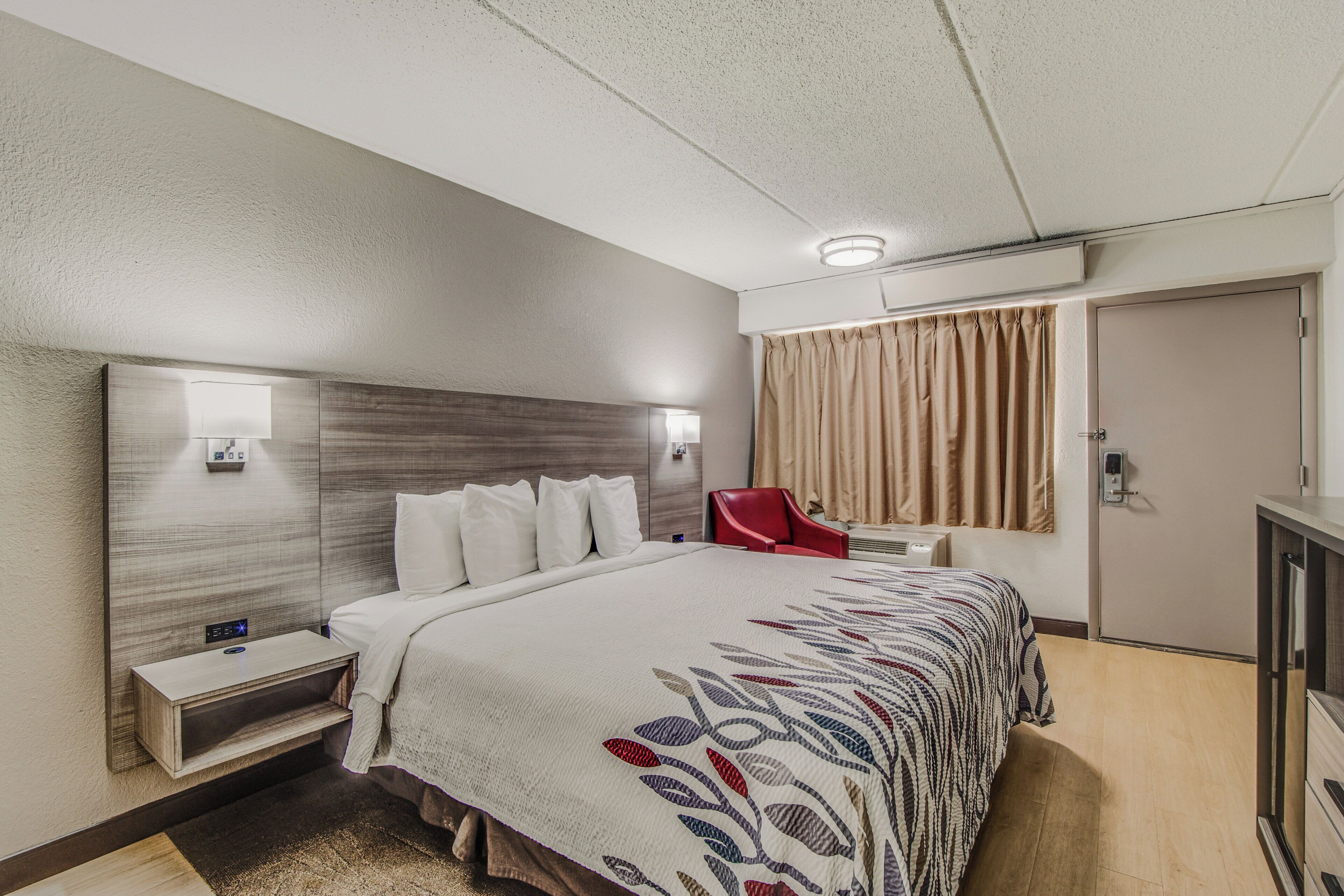 Quarto de hotel com cama, uma cadeira vermelha, escrivaninha e guarda-roupa.