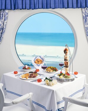Mesa de jantar com vista para a praia, diversos pratos e uma taça de vinho.