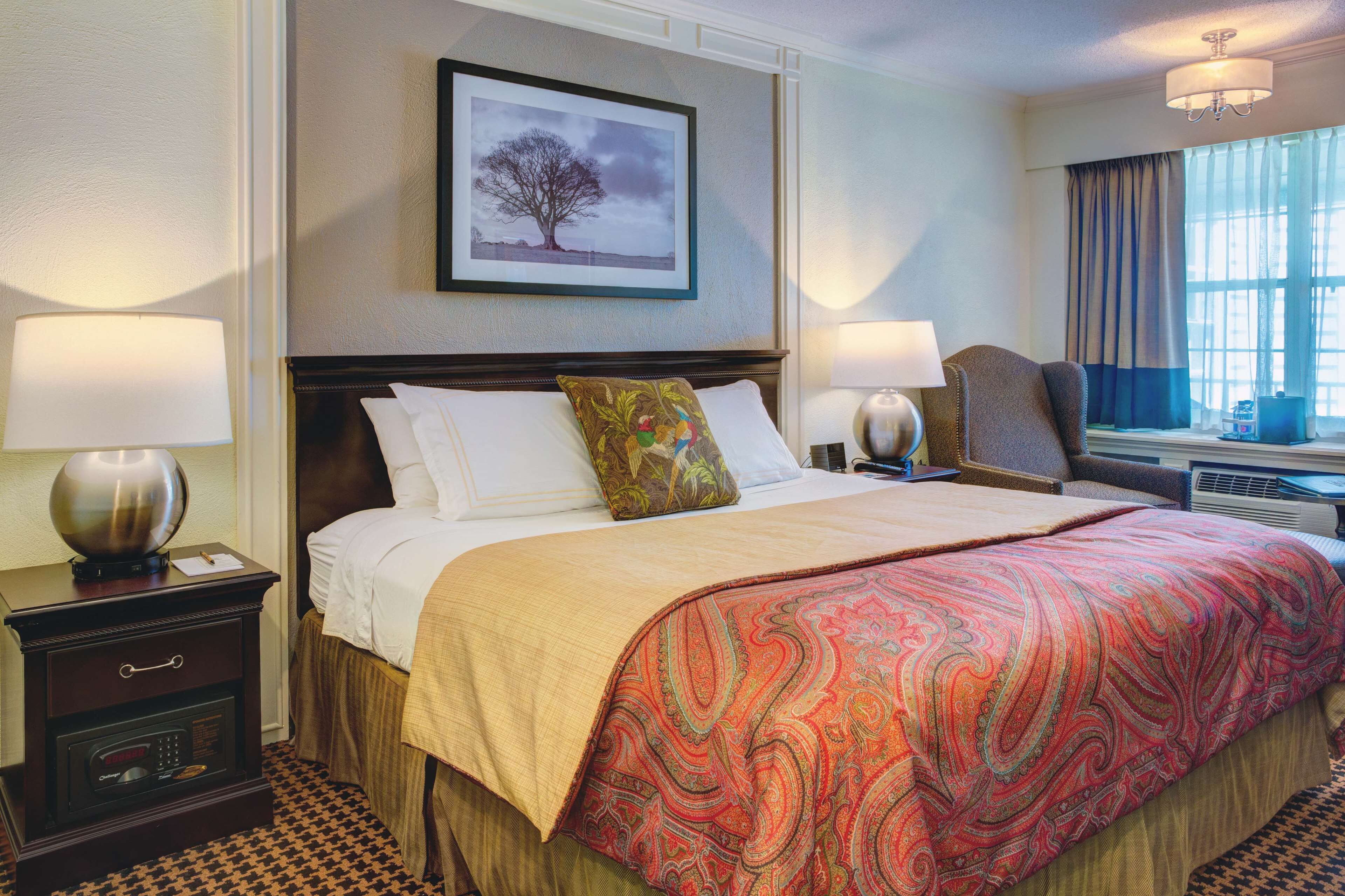 Quarto de hotel com uma cama grande, duas mesas de cabeceira, uma cadeira, uma janela com cortinas e um quadro na parede.