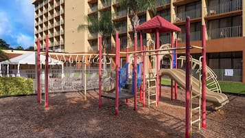 Parquinho infantil com escorrega e estruturas de escalada, cercado por um edifício com sacadas.