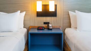 Quarto de hotel com duas camas, mesa de cabeceira com relógio e telefone, e luminária fixada na parede.