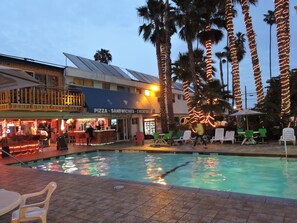 Uma área de piscina com espreguiçadeiras e bar, cercada por palmeiras e um edifício com placa de pizza e sanduíches.