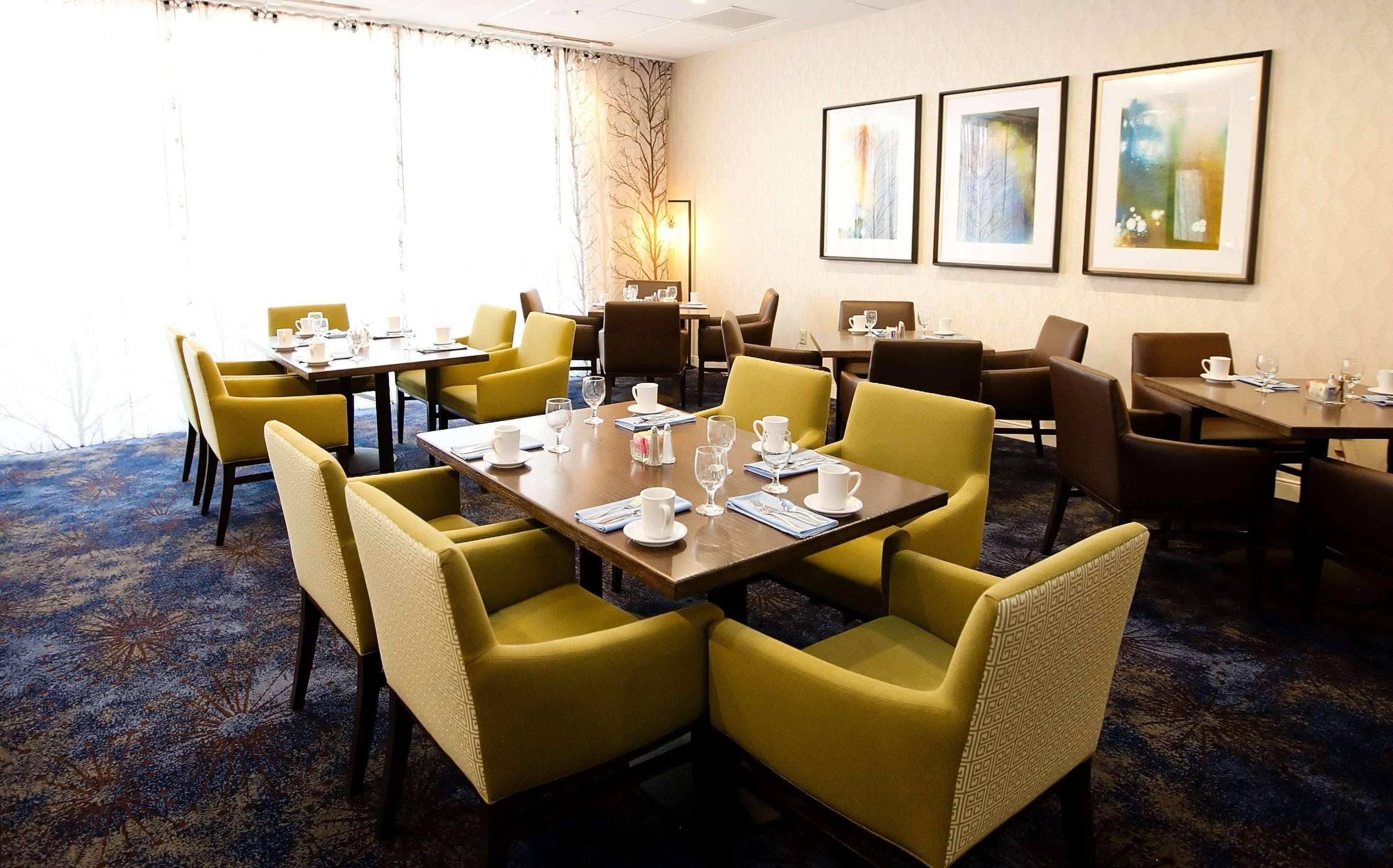 Uma sala de jantar de hotel com mesas preparadas para quatro pessoas, cadeiras amarelas e obras de arte emolduradas na parede.
