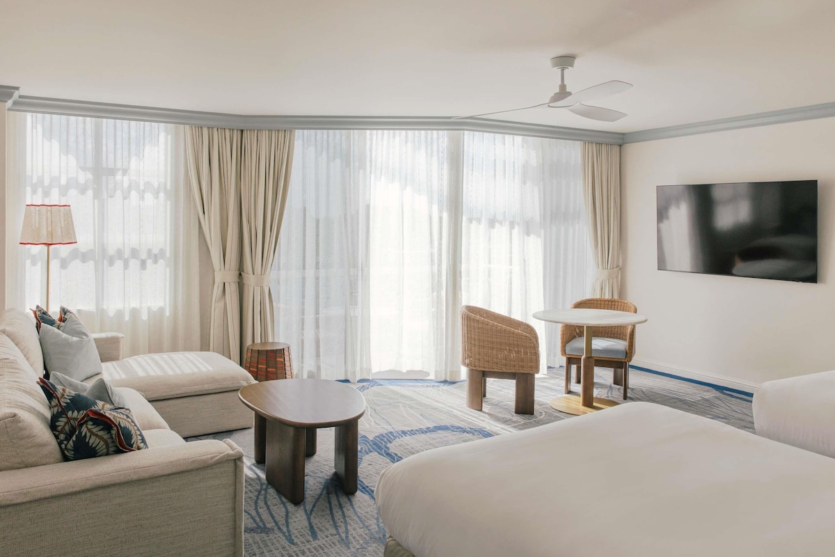 Quarto de hotel com cama, sofá, poltrona, mesa pequena e televisão na parede.