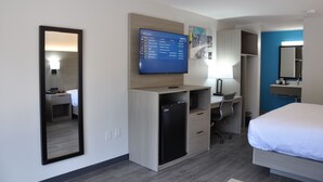 Quarto de hotel com cama, escrivaninha, cadeira, televisão, espelho e guarda-roupa.
