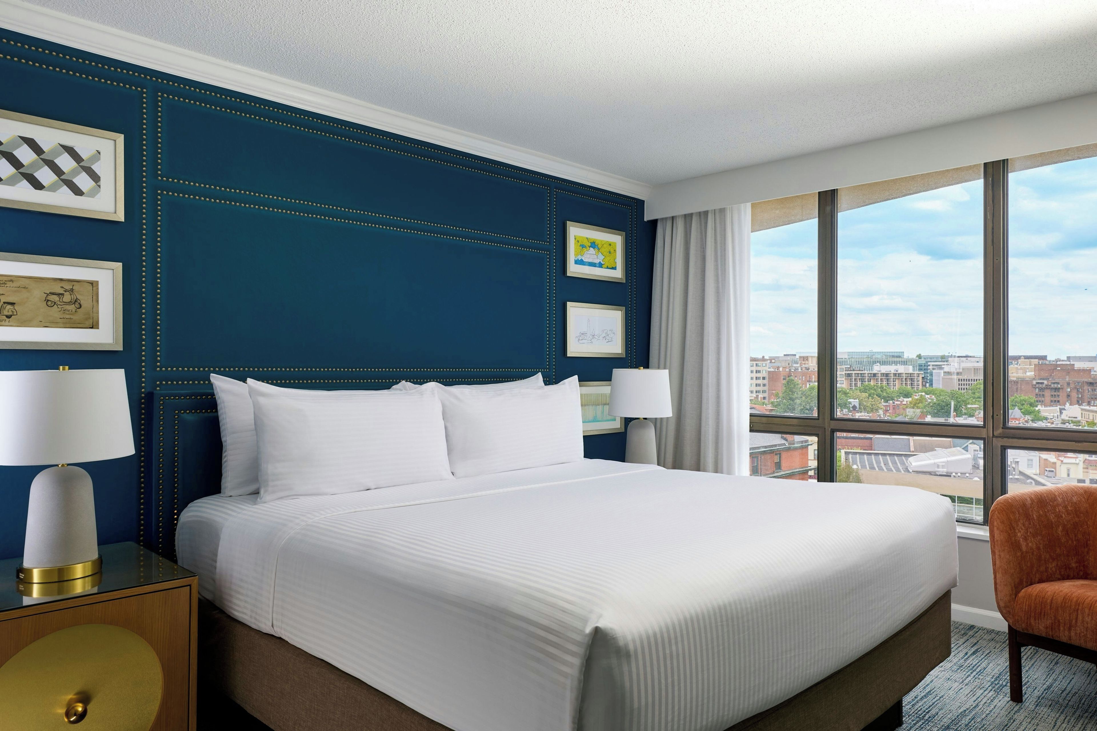 Quarto de hotel com uma cama grande, mesa de cabeceira, luminária, poltrona e vista para a cidade pela janela.