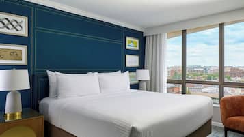Quarto de hotel com uma cama grande, mesa de cabeceira, luminária, poltrona e vista para a cidade pela janela.