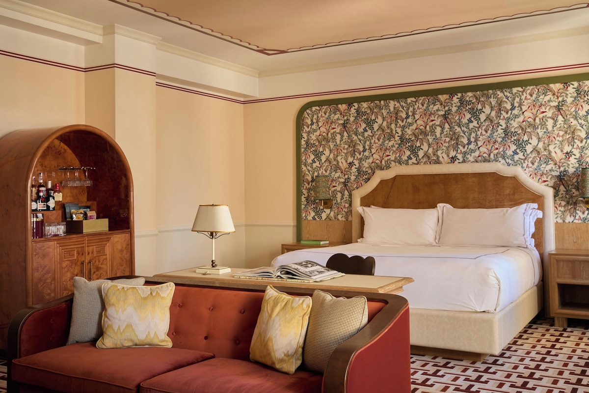 Quarto de hotel com cama, sofá, aparador de madeira com garrafas e papel de parede estampado.