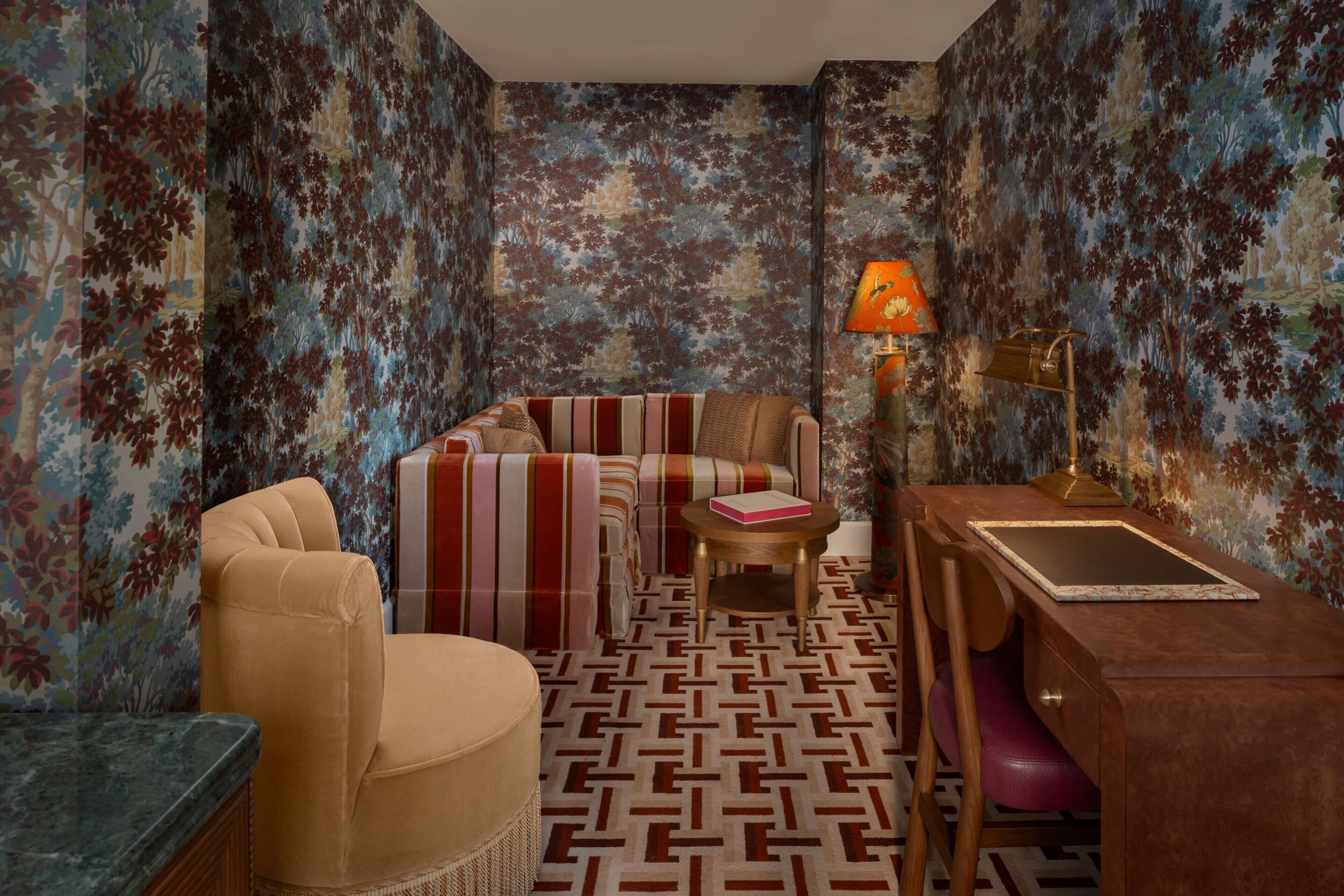 Um quarto com carpete estampado, um sofá, uma poltrona, uma escrivaninha com abajur e um papel de parede floral.