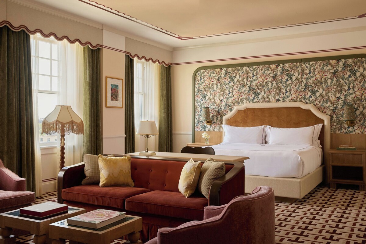 Quarto de hotel com uma cama grande, um sofá, uma escrivaninha com abajur e papel de parede estampado.