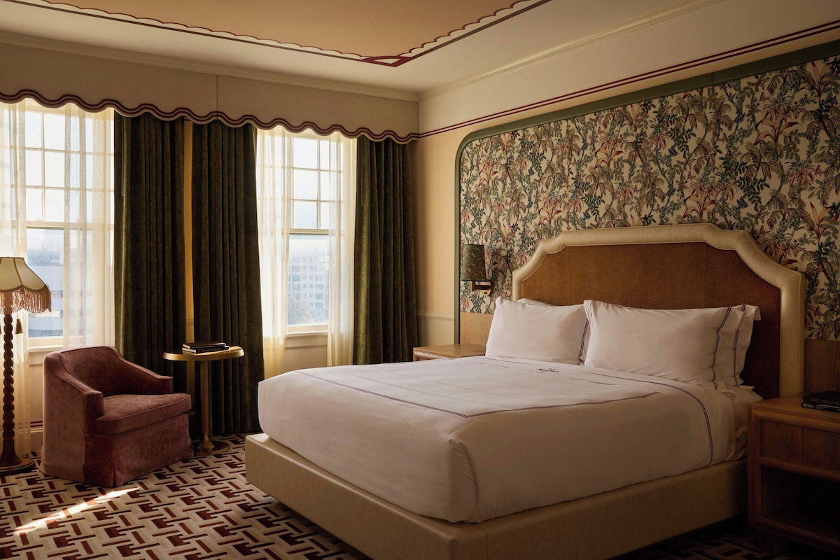 Quarto de hotel com uma cama grande, uma cadeira, uma mesinha e uma luminária.