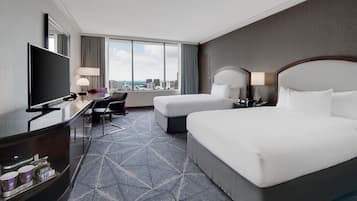 Quarto de hotel com cama, escrivaninha com televisão, cadeira, janela e vista para a cidade, além de carpete.
