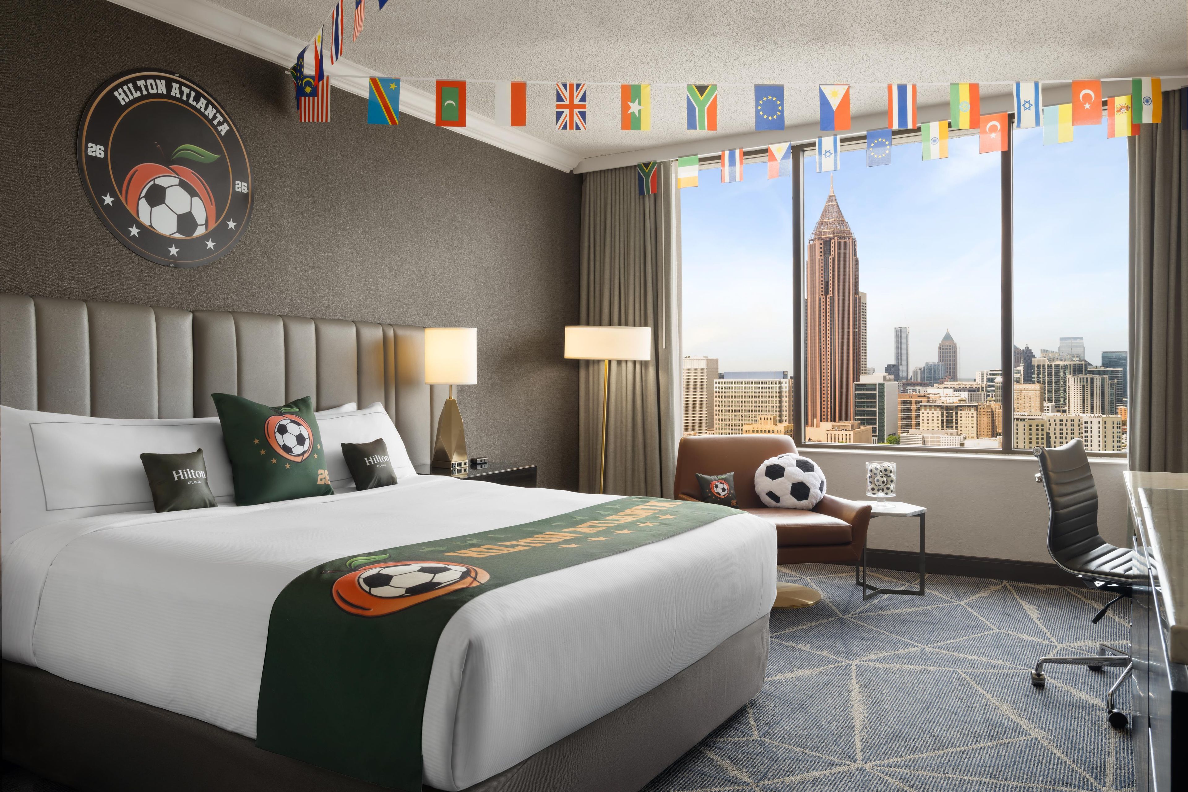 Quarto de hotel com uma cama grande, uma escrivaninha, uma cadeira e vista para o horizonte da cidade.