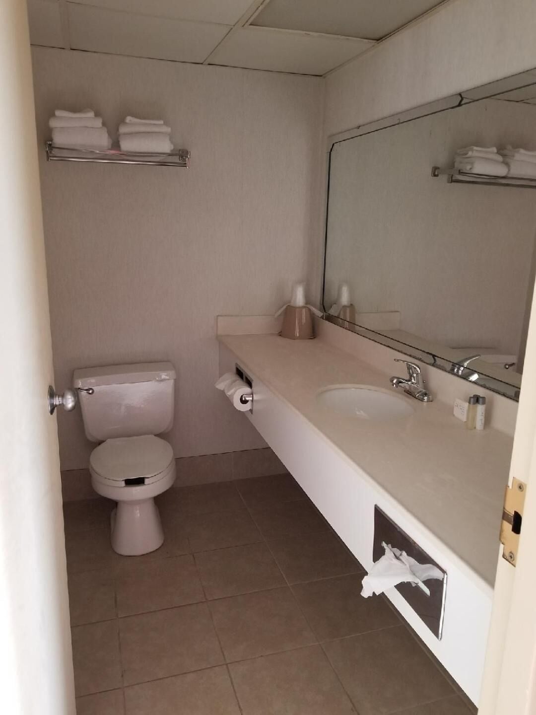 Uma casa de banho pequena e limpa, com sanita, lavatório e espelho. Há prateleiras sobre a sanita com toalhas.