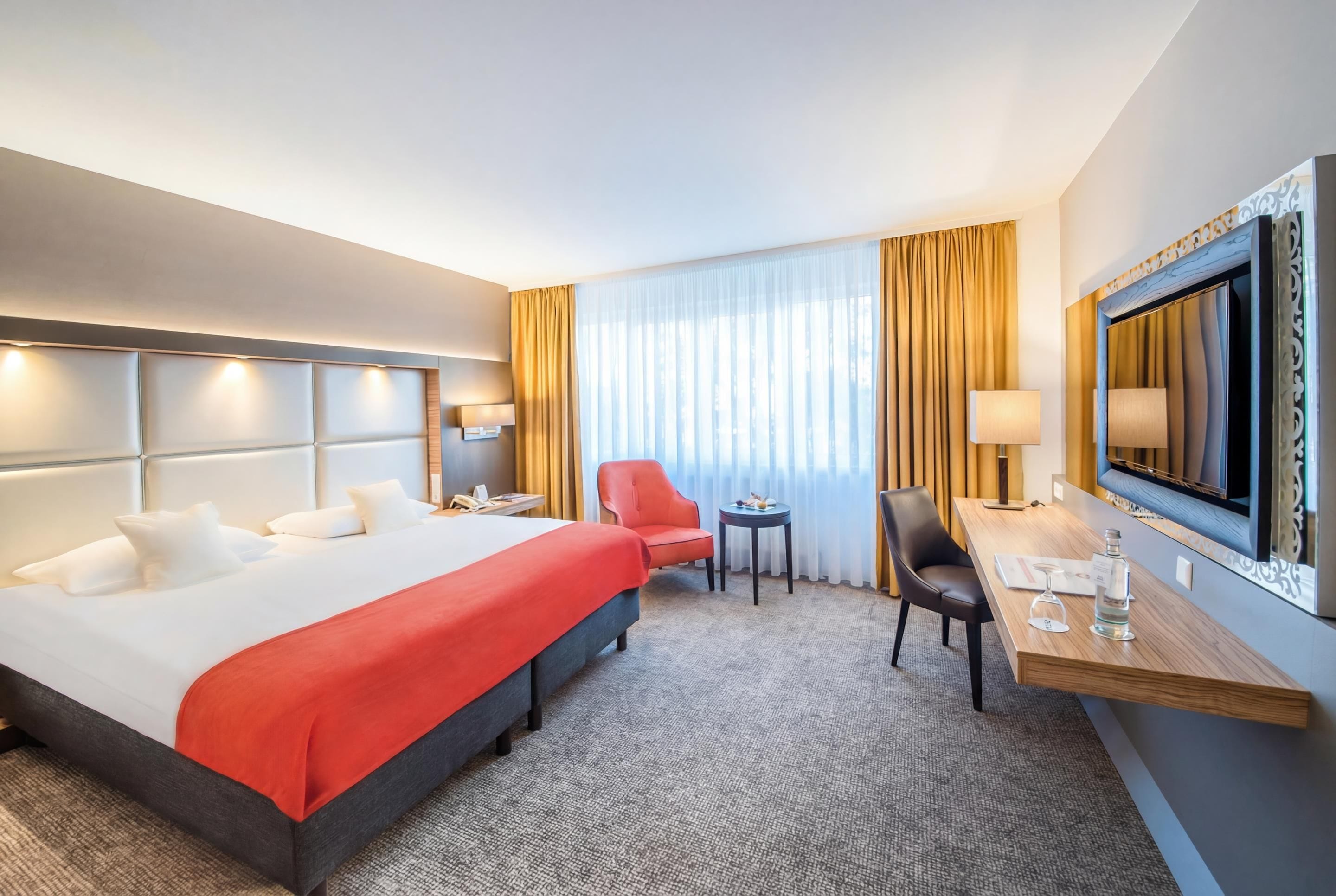 Quarto de hotel com cama, escrivaninha, cadeira, televisão e janela com cortinas.
