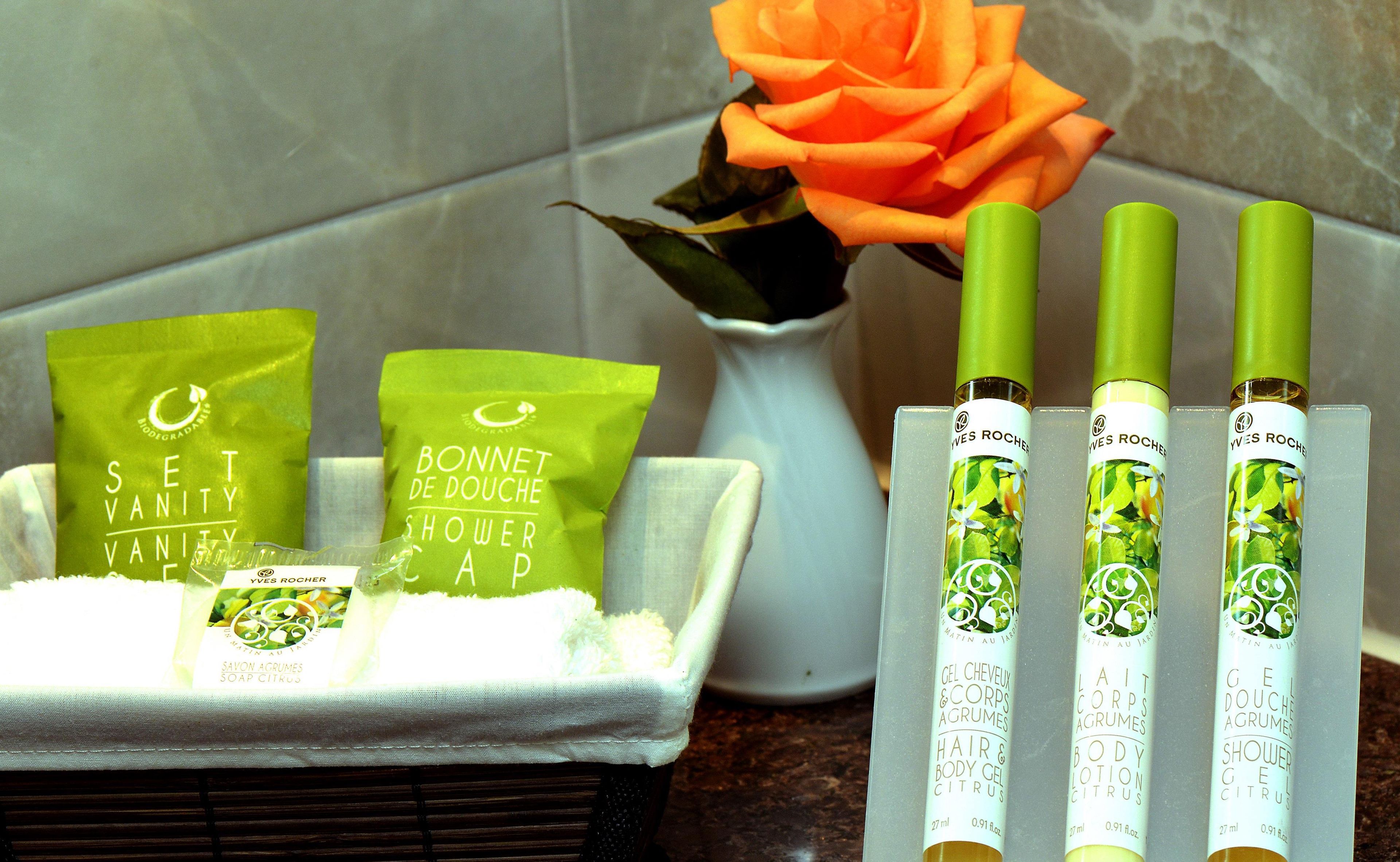Uma toucador de banheiro com produtos de higiene pessoal, incluindo um touca de banho verde, um vaso branco com uma rosa laranja e quatro frascos de loção corporal Yves Rocher.