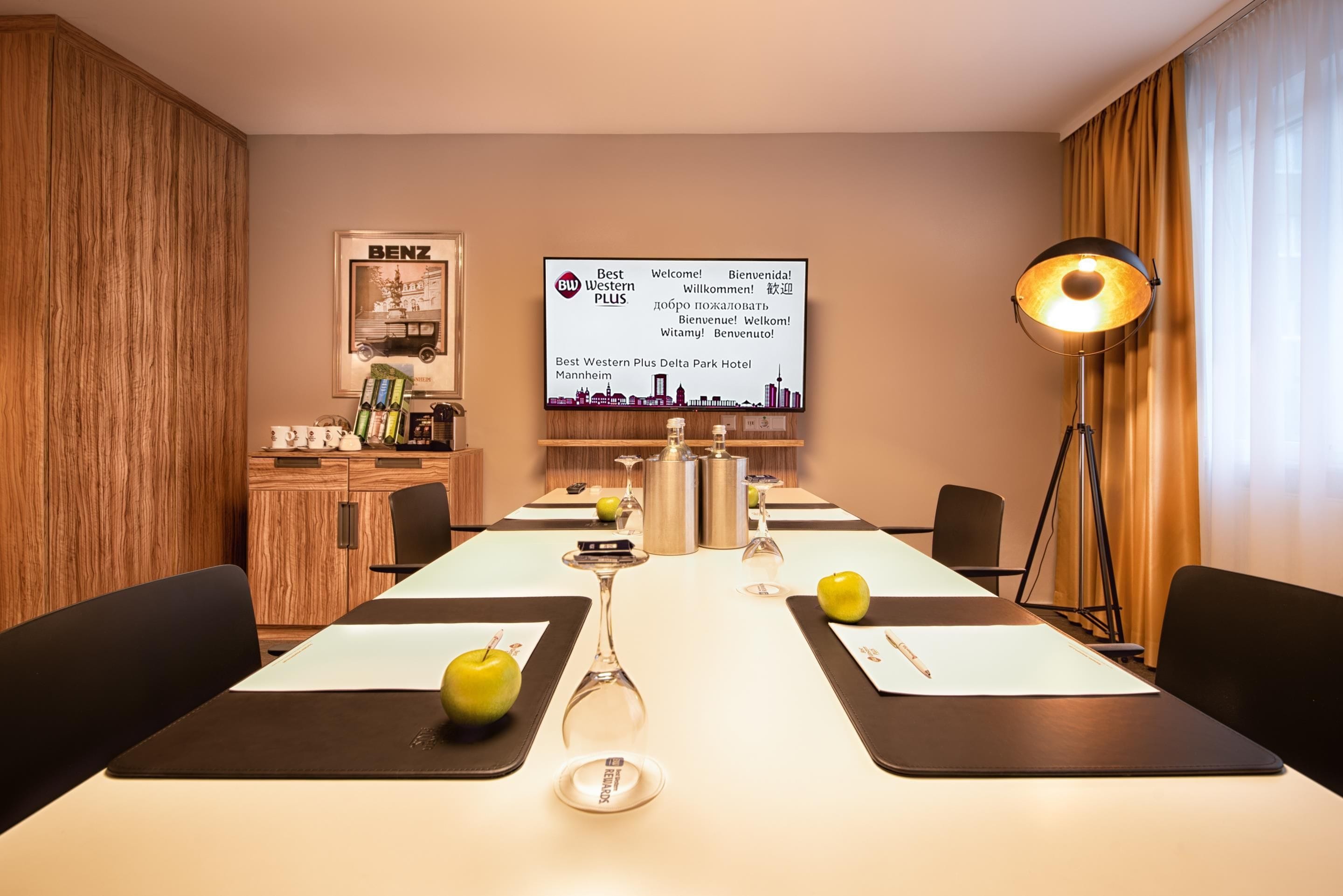Sala de conferência com uma mesa comprida, cadeiras, relógio, placa de boas-vindas emoldurada e abajur.