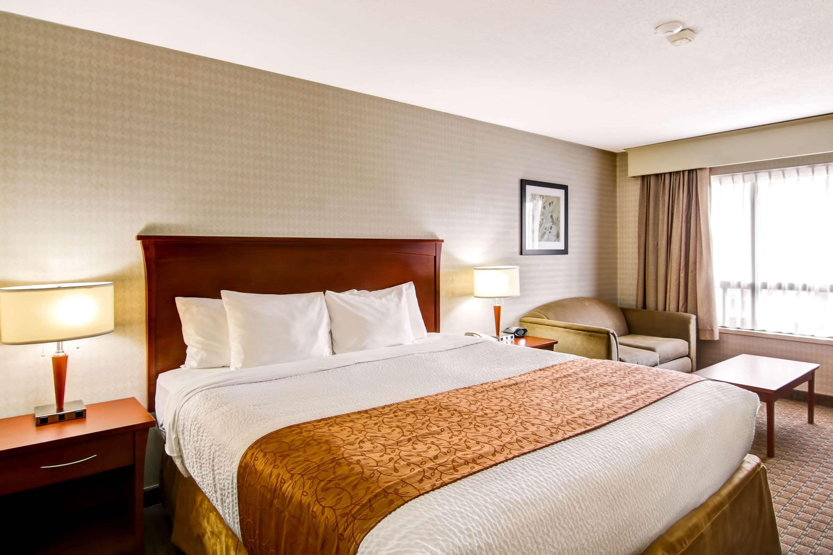 Quarto de hotel com uma cama grande, duas mesas de cabeceira com abajures, uma cadeira, uma pequena mesa e uma janela com cortinas.