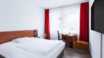 Quarto de hotel com uma cama grande, uma escrivaninha com cadeira, uma televisão e uma luminária.