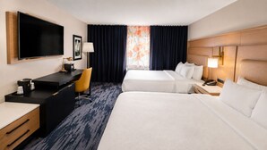 Quarto de hotel com televisão de tela plana, escrivaninha com cadeira, duas camas, mesa de cabeceira e janela com cortinas.
