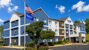 Um hotel Fairfield by Marriott com um mastro de bandeira, estacionamento e área ajardinada com arbustos e flores.