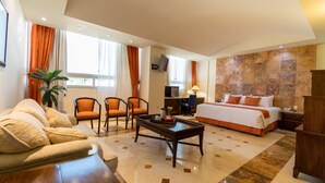 Quarto de hotel com cama, área de estar com cadeiras, mesa de centro, televisão e uma grande janela com cortinas.